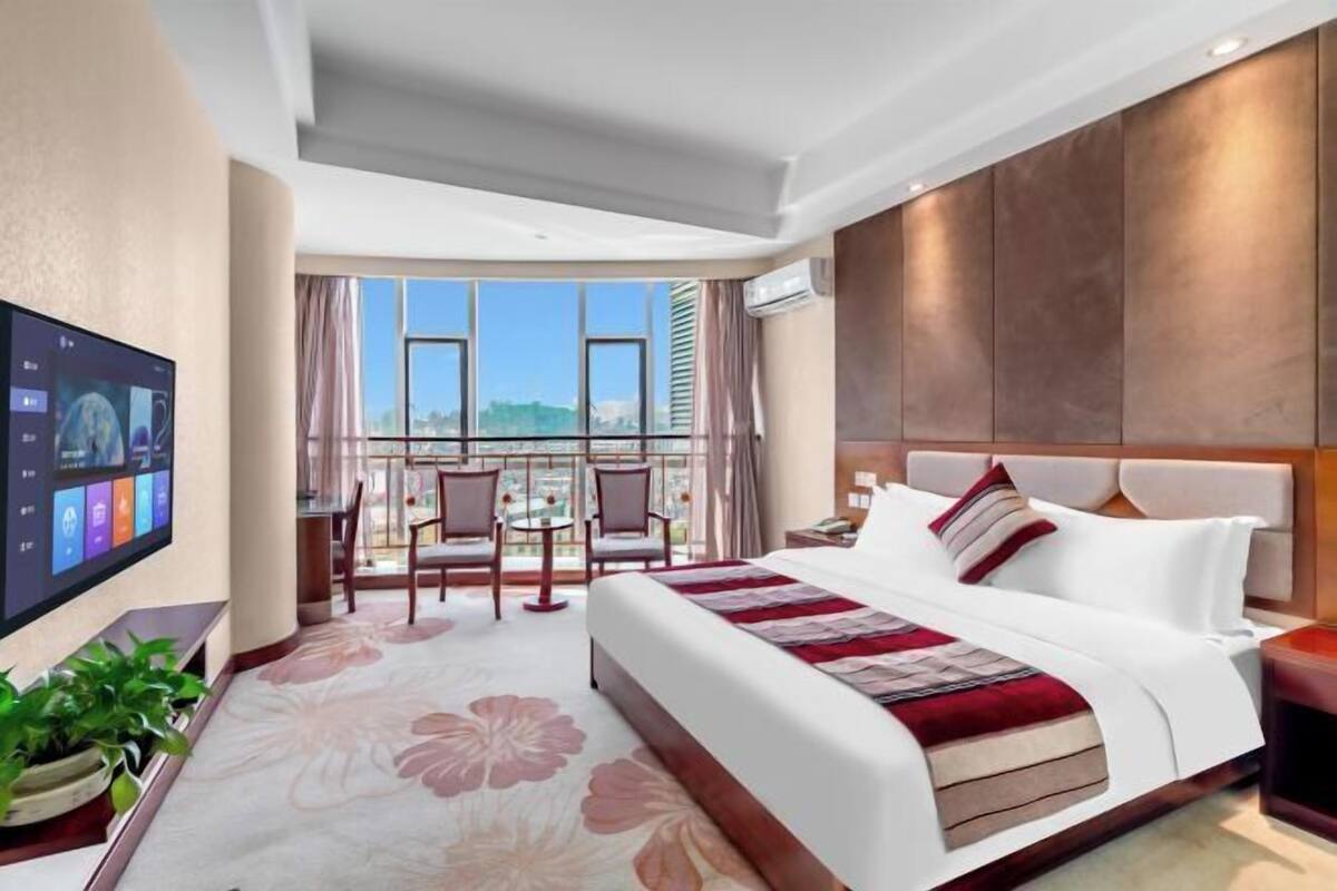 Quarto de hotel com uma cama grande, televisão, escrivaninha com cadeiras e vista para o exterior.