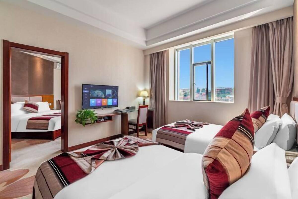 Quarto de hotel com duas camas, televisão, escrivaninha e uma janela grande com cortinas.