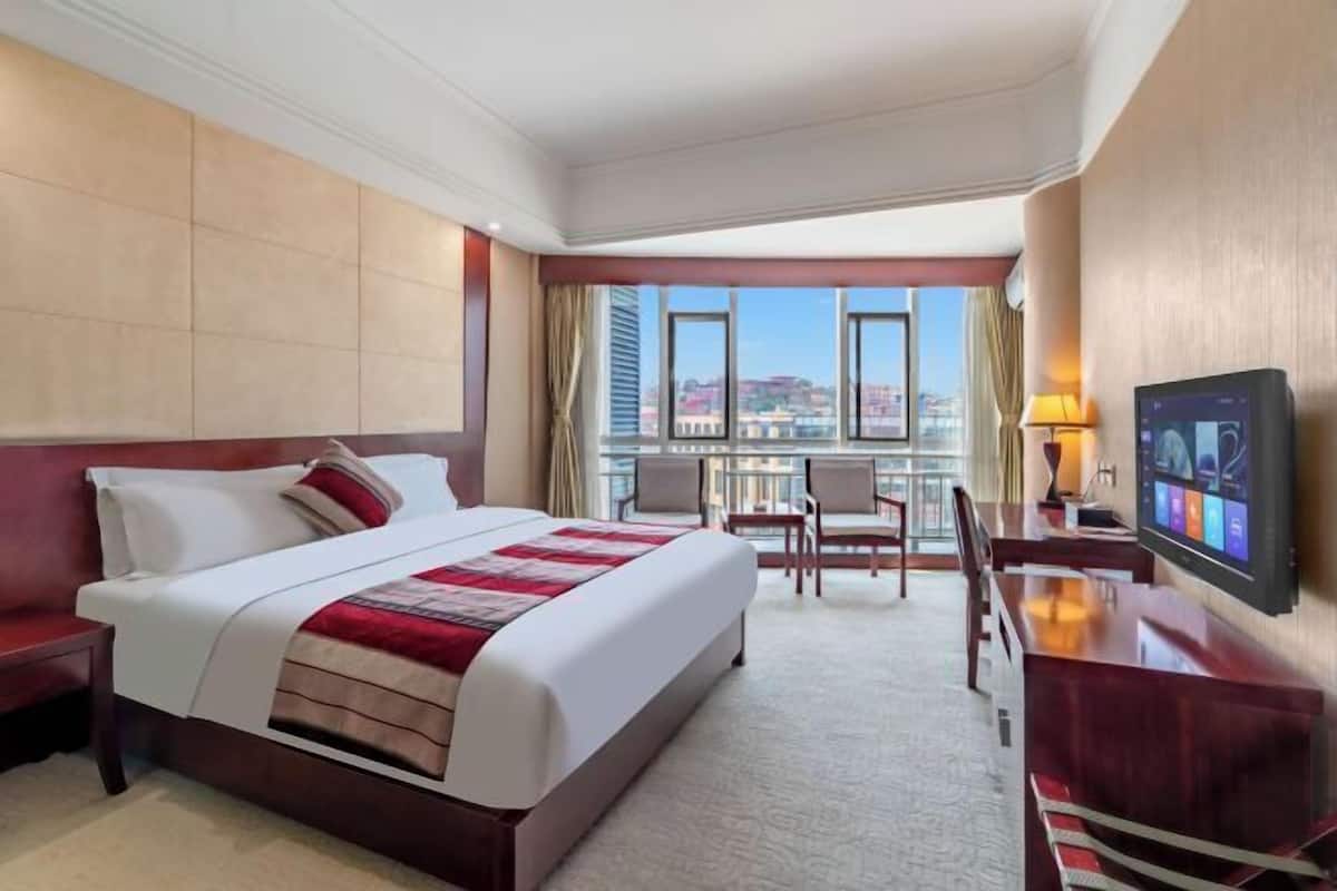 Quarto de hotel com uma cama grande, uma mesa de trabalho com televisão e uma varanda com vista para a cidade.