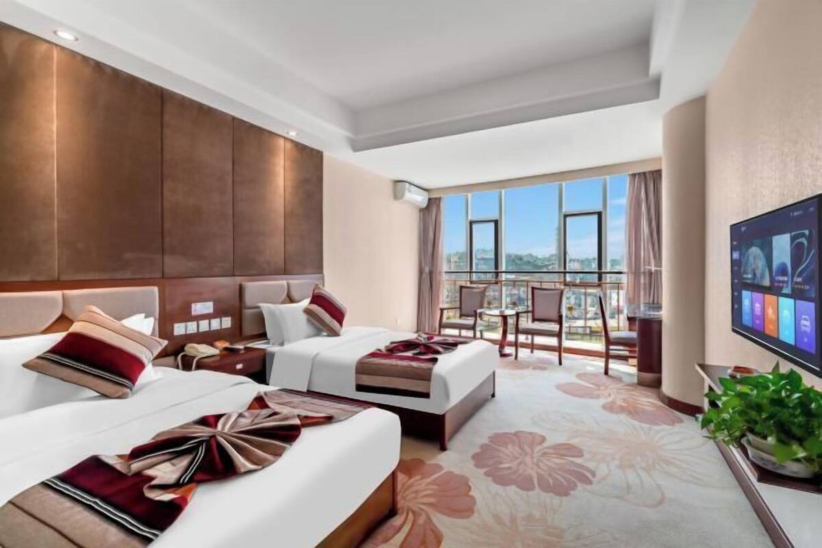 Quarto de hotel com duas camas, varanda com mesa e cadeiras, televisão e uma janela grande.
