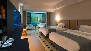 Quarto de hotel com duas camas, uma escrivaninha com cadeira, uma janela grande e um minibar.
