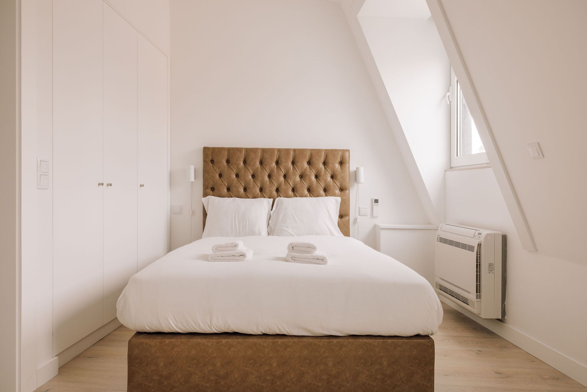 Quarto com cabeceira estofada, roupa de cama branca, um banco de madeira e ar condicionado instalado.