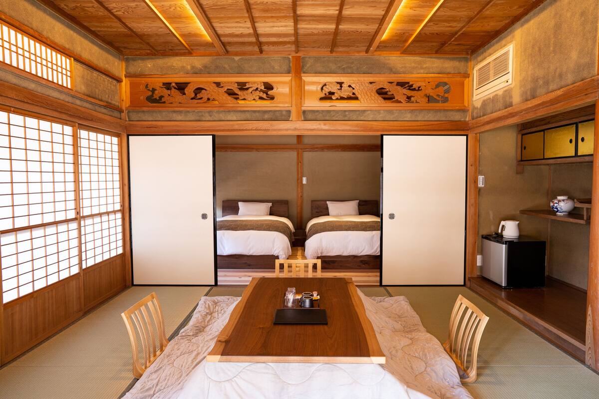 Um quarto tradicional japonês com tatames, uma mesa de jantar de madeira, duas camas com roupa de cama branca e uma pequena área de cozinha com chaleira e bule.