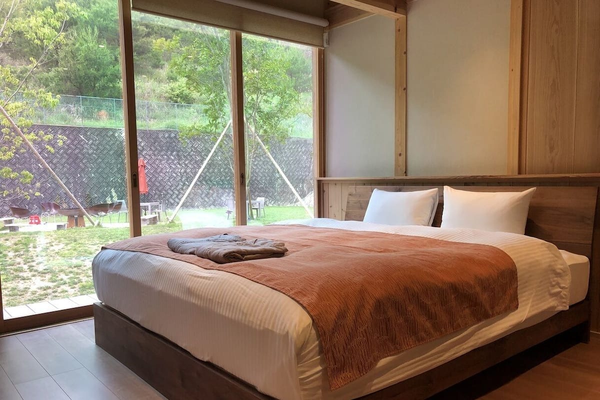 Quarto com cama grande, cabeceira de madeira e vista para o exterior através de uma janela ampla.