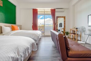 Quarto de hotel com duas camas, um sofá, uma varanda com vista, uma escrivaninha e um ventilador.