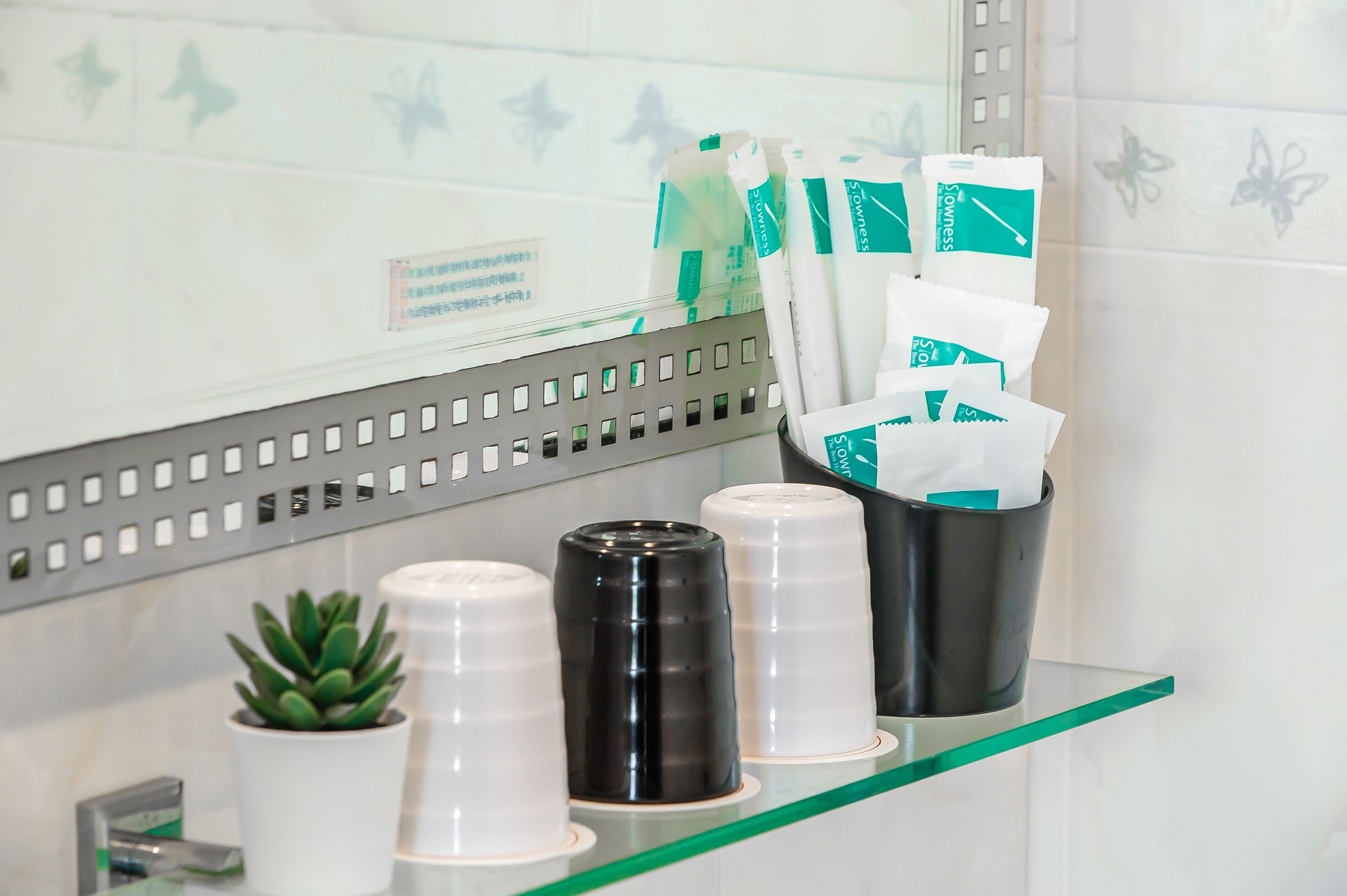 Casa de banho com prateleira de vidro, vários produtos de higiene pessoal, uma planta em vaso e espelho com desenhos de borboletas.