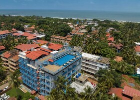 Hotel com vários andares e piscina no terraço, cercado por edifícios residenciais e palmeiras, com vista para uma praia.