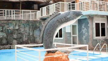 Uma grande escultura de peixe em uma piscina, com guarda-corpo branco e parede de pedra ao fundo.