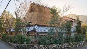 Uma casa japonesa tradicional com telhado de palha, cercada por um muro de pedra e vegetação exuberante.