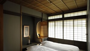 Quarto em estilo japonês tradicional, com piso de tatame, teto de madeira baixo e janela com tela shoji. Há duas camas com roupa de cama branca, uma mesa pequena com um abajur e um quadro emoldurado.