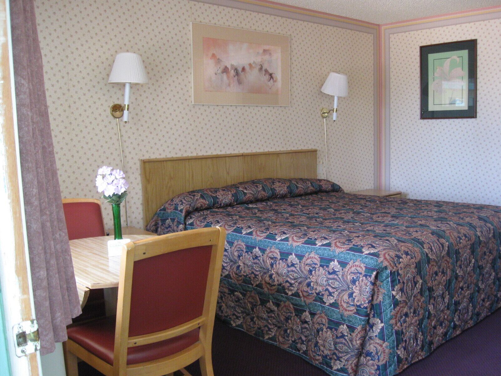 Quarto de hotel com uma cama, duas luminárias de cabeceira, um quadro na parede, uma mesinha com um vaso de flores e uma cadeira.