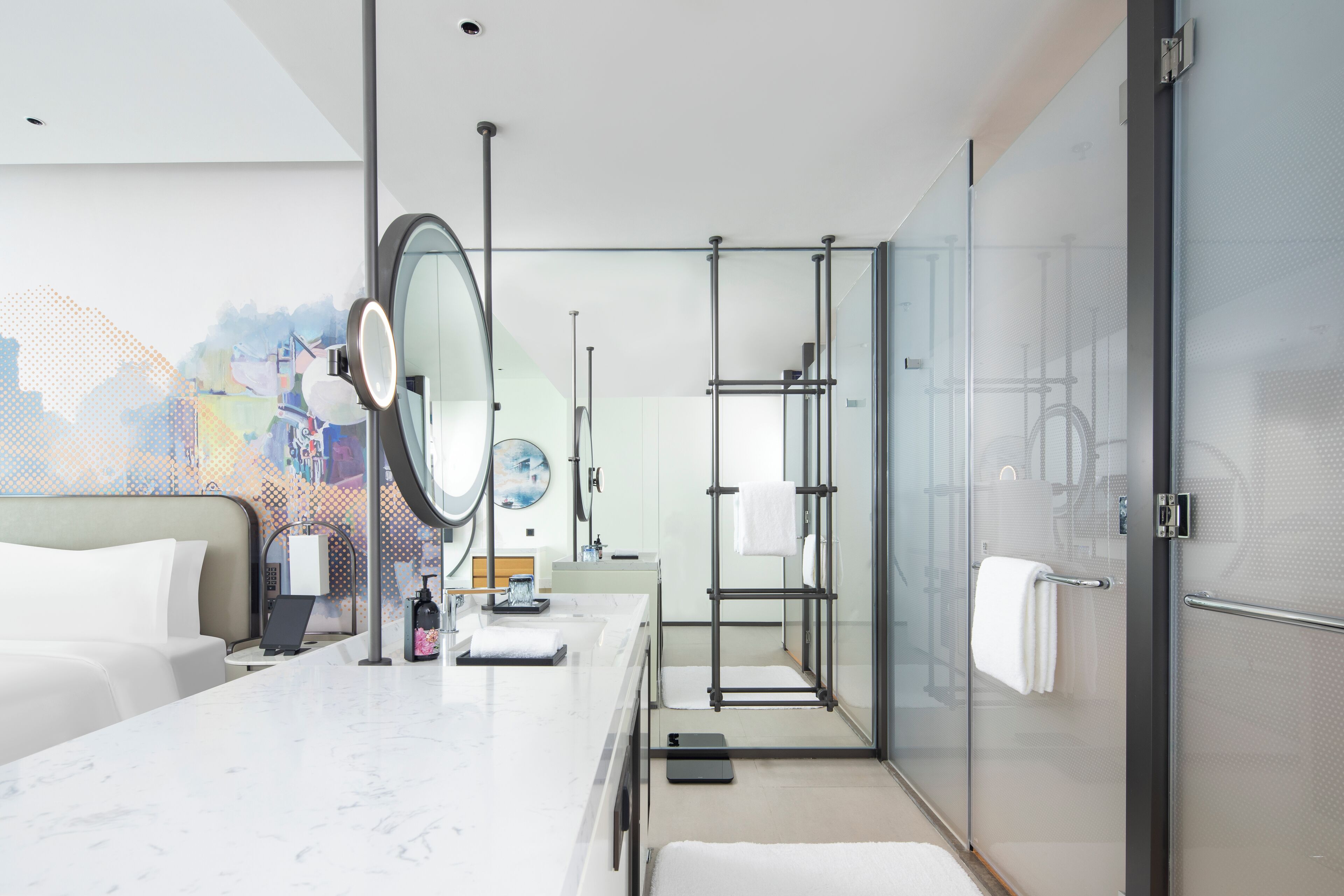 Quarto de hotel moderno com uma cama grande, toucador com bancada de mármore, box de vidro para o chuveiro e um mural com vista para a cidade na parede.