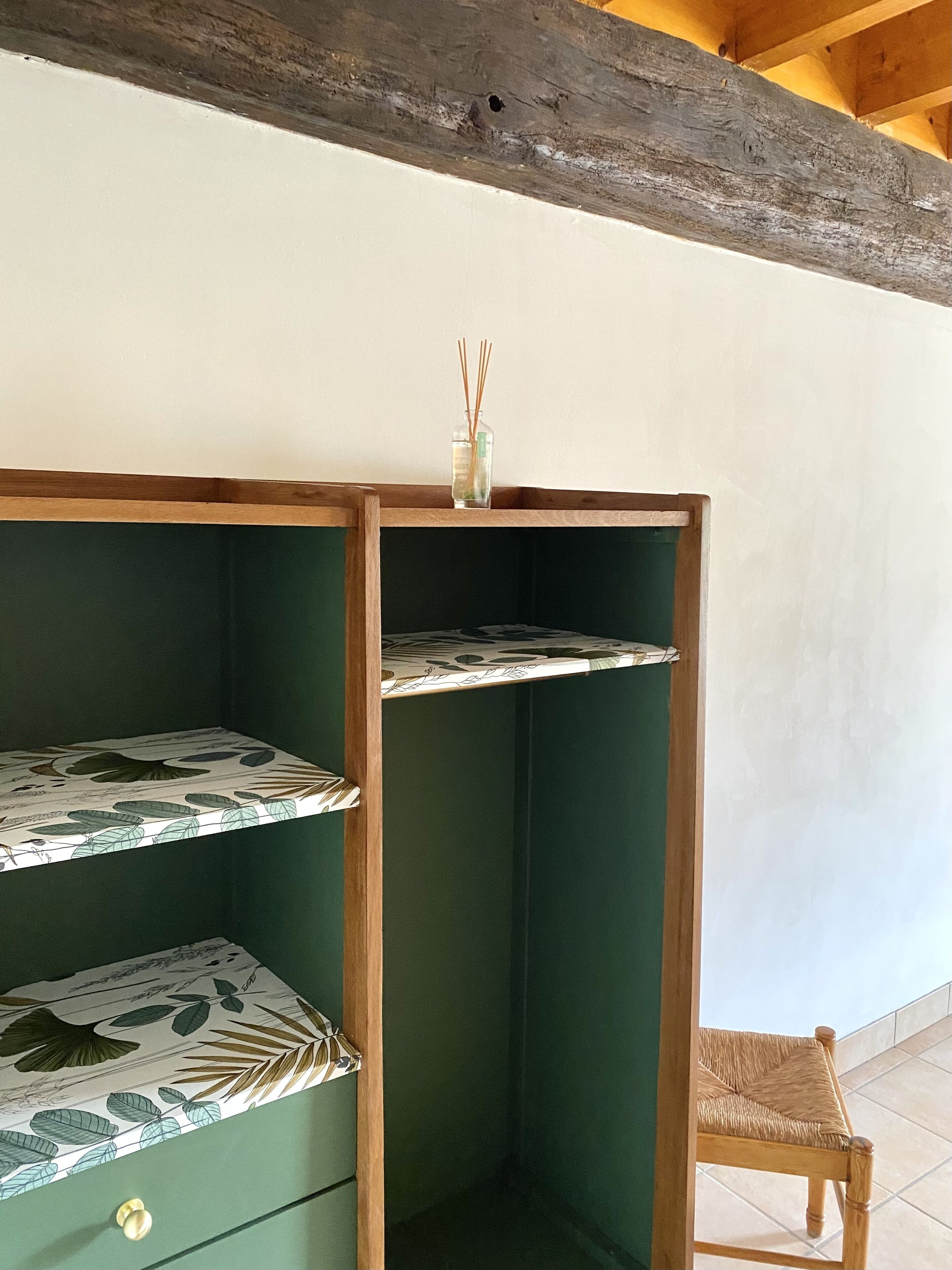 Canto de um quarto com um armário verde, uma cadeira de madeira pequena e um frasco de varetas em uma estante.