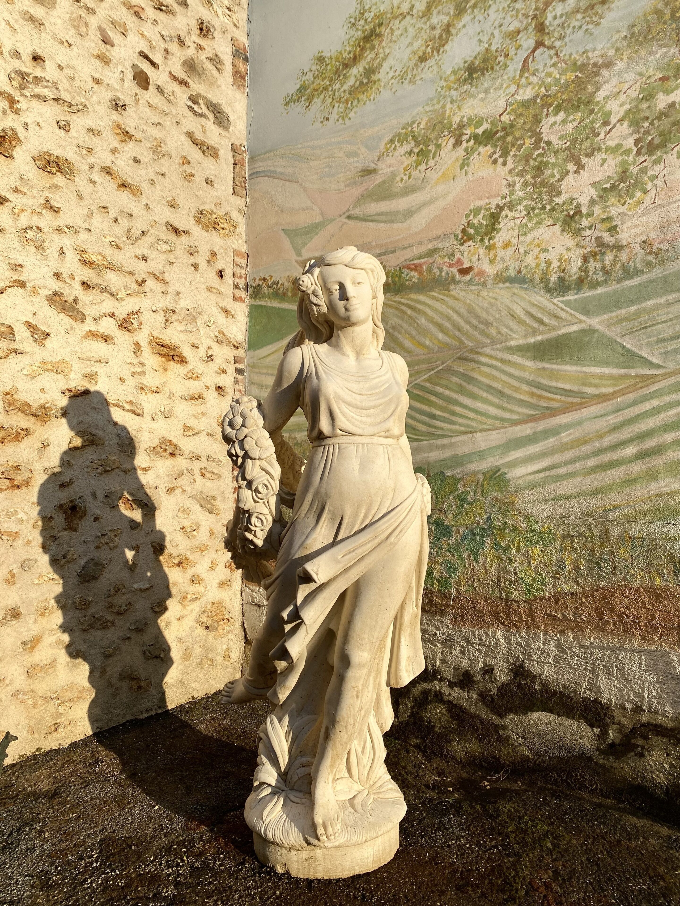 Estátua de uma mulher vestida em estilo clássico, posicionada diante de uma parede texturizada com um mural de paisagem ao fundo.