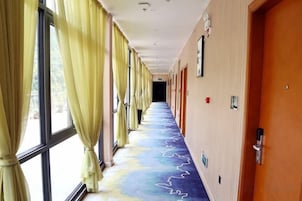 Corredor de hotel com carpete estampado, janelas amplas com cortinas e portas à direita.