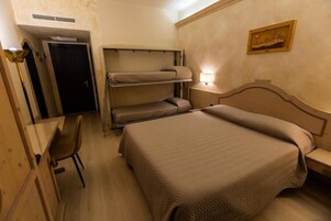 Quarto de hotel com uma cama de solteiro, uma escrivaninha com cadeira, uma mesinha de cabeceira e uma luminária fixada na parede.