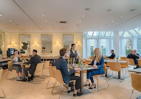 Uma sala de jantar moderna com várias mesas e cadeiras, um garçom atendendo aos hóspedes e grandes janelas que proporcionam luz natural.