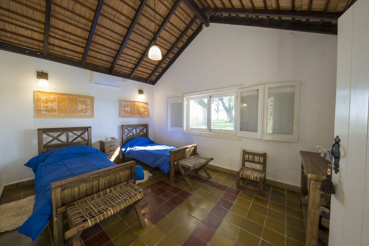 Quarto com duas camas, um banco, uma mesa pequena e uma janela.