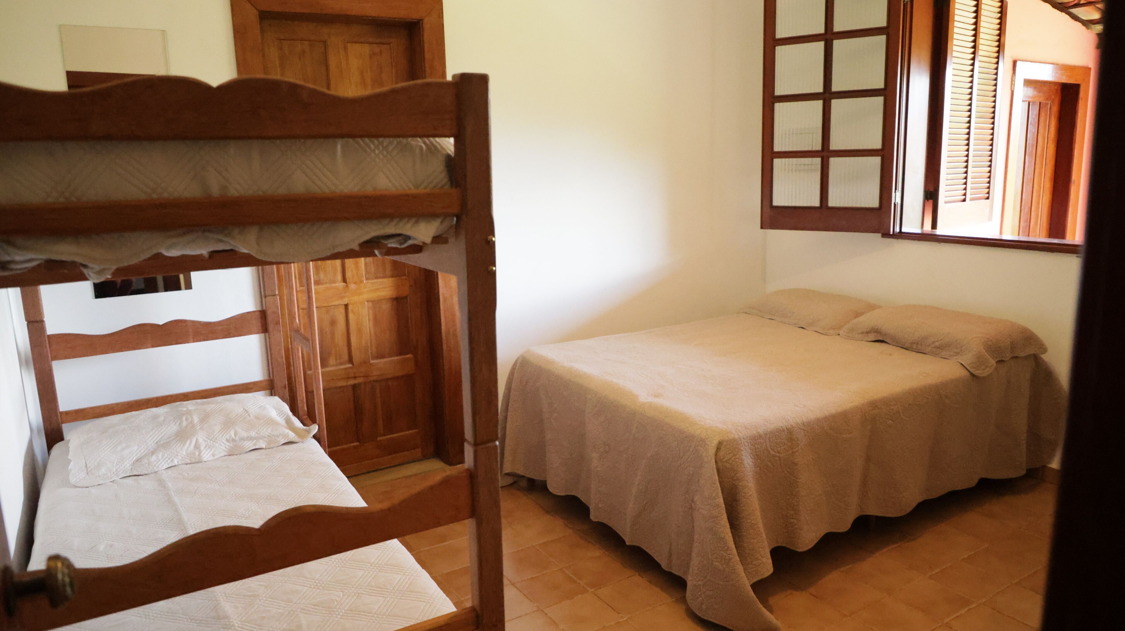 Quarto com cama de solteiro e uma cama individual. Há uma porta de madeira e uma janela com persianas.