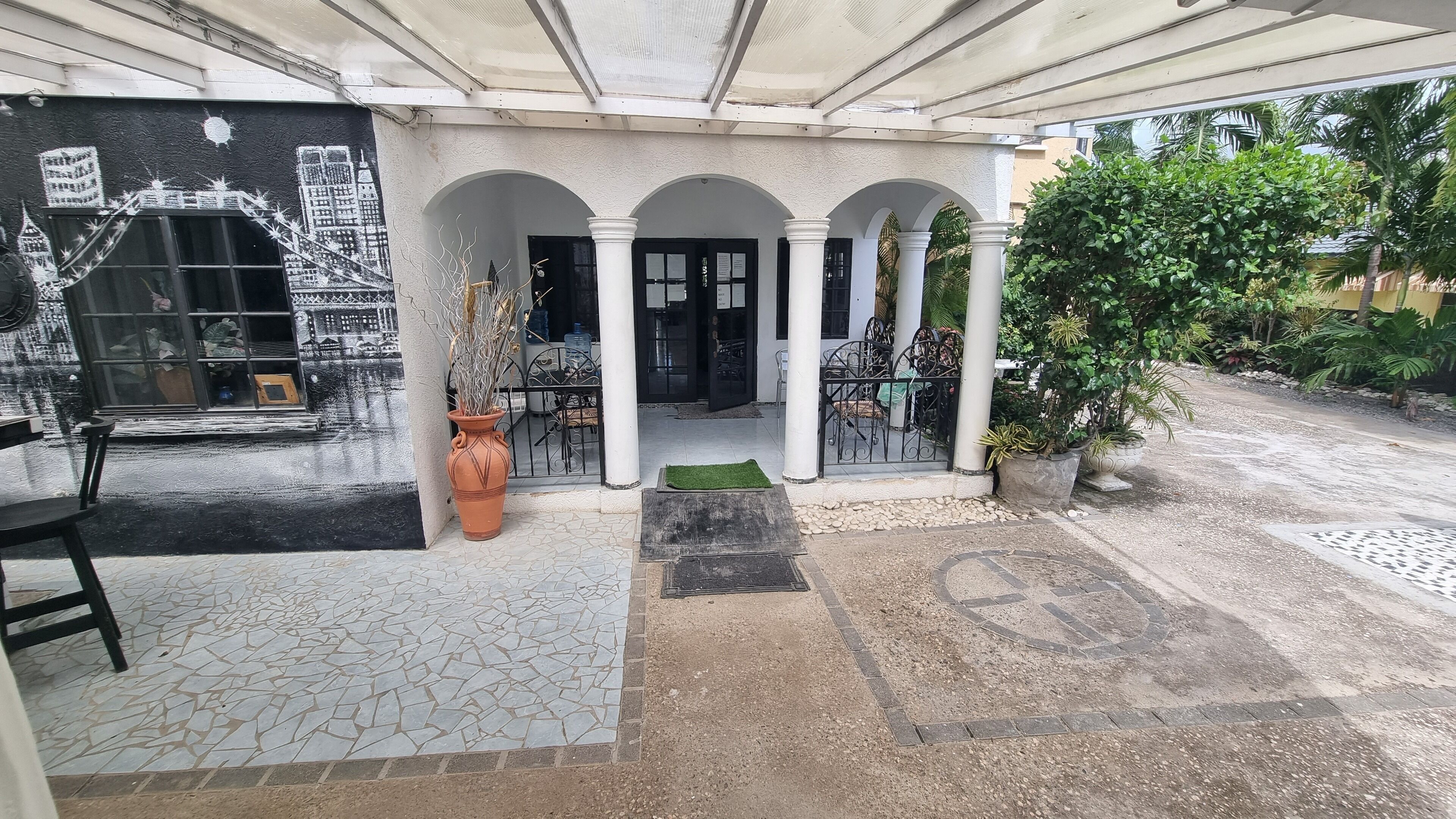 Um espaço ao ar livre coberto, com piso de azulejos, uma grande planta em vaso e um mural na parede.