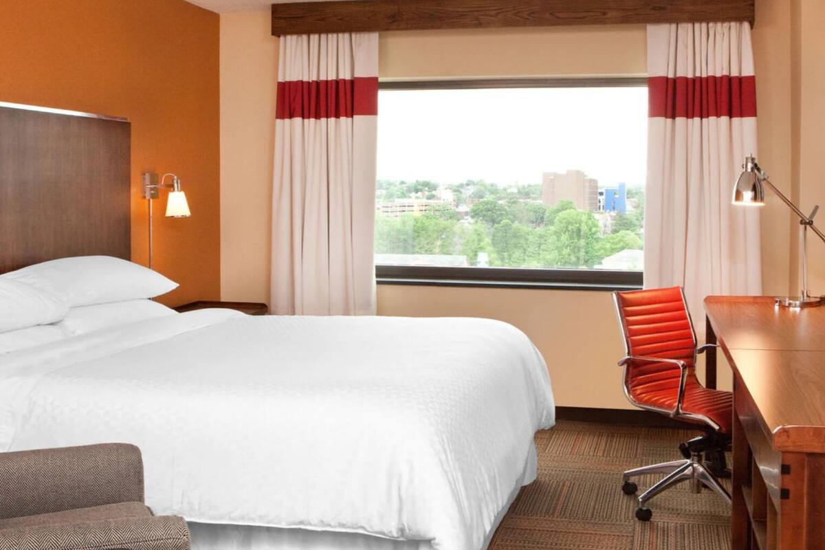 Quarto de hotel com uma cama grande, uma escrivaninha com cadeira, televisão e janela com cortinas.