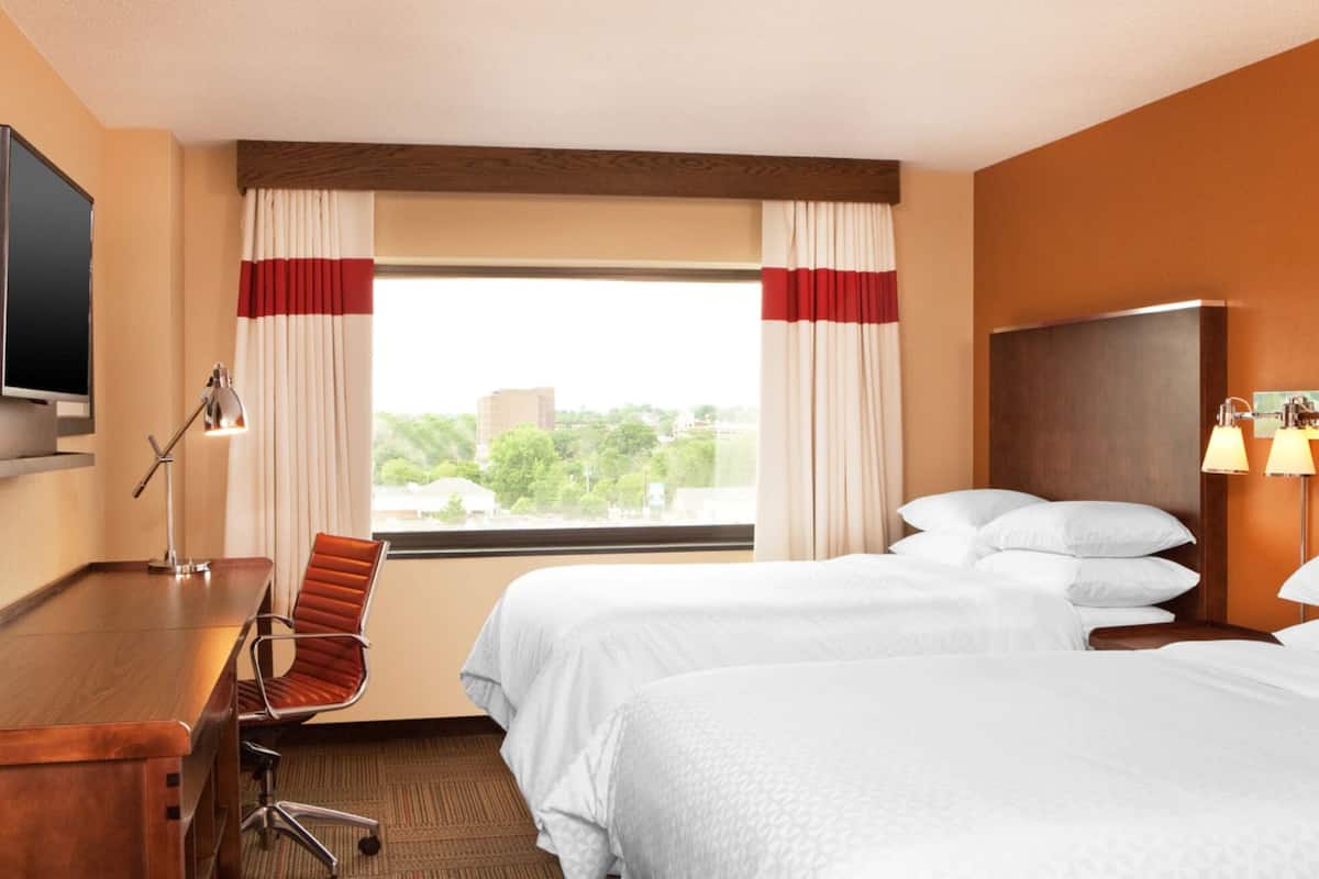 Quarto de hotel com duas camas, uma escrivaninha com cadeira, televisão de tela plana na parede e uma janela com cortinas.