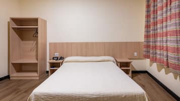Quarto de hotel com uma cama, um guarda-roupa de madeira, uma mesa de cabeceira com telefone e uma cortina listrada.