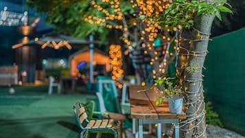 Uma área externa com mesa e cadeiras de madeira, luzes de corda e uma árvore com plantas trepadeiras.