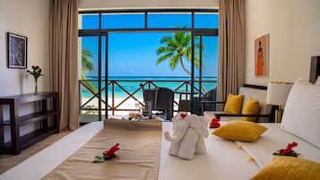 Quarto de hotel com uma grande janela com vista para uma praia e oceano, uma cama com roupa de cama branca, uma mesinha com um copo de vinho e uma figura decorativa de elefante.