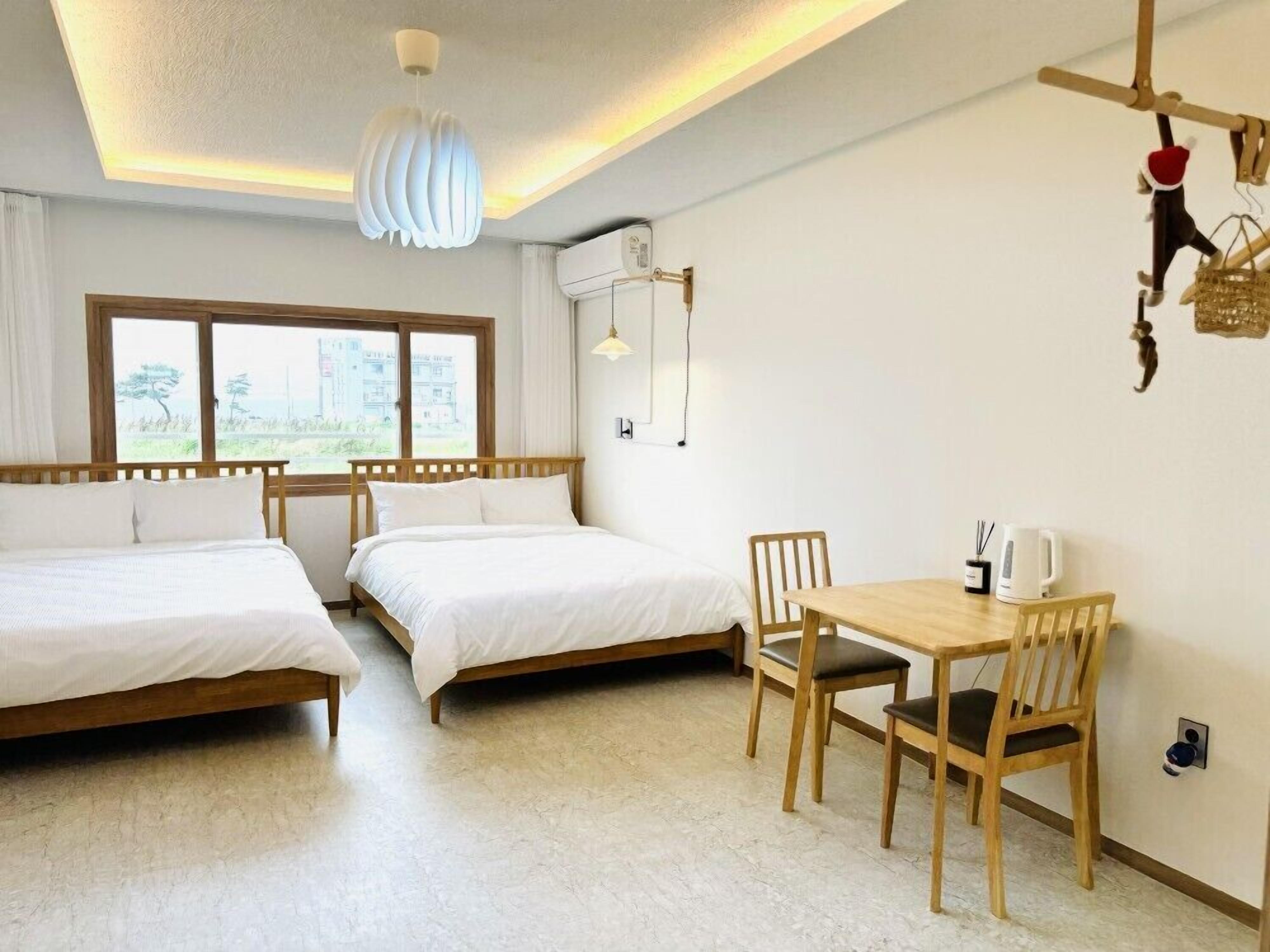Quarto de hotel com duas camas de solteiro, uma mesa de madeira com cadeiras, luminária pendente e vista para o exterior através de amplas janelas.