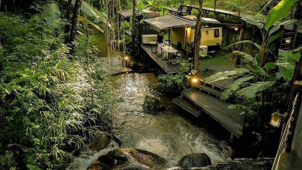 Um hotel de estilo cabana rústica, com um pequeno riacho atravessando a propriedade, rodeado por vegetação tropical densa.