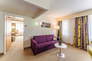 Quarto de hotel com sofá roxo, uma mesa redonda pequena com uma garrafa e copos, uma luminária e um quadro na parede.