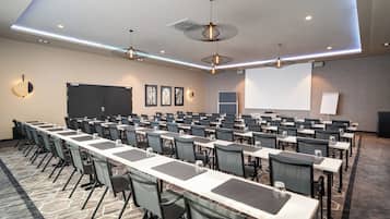 Uma sala de conferências com mesas compridas e cadeiras, uma tela de projetor e um quadro branco.