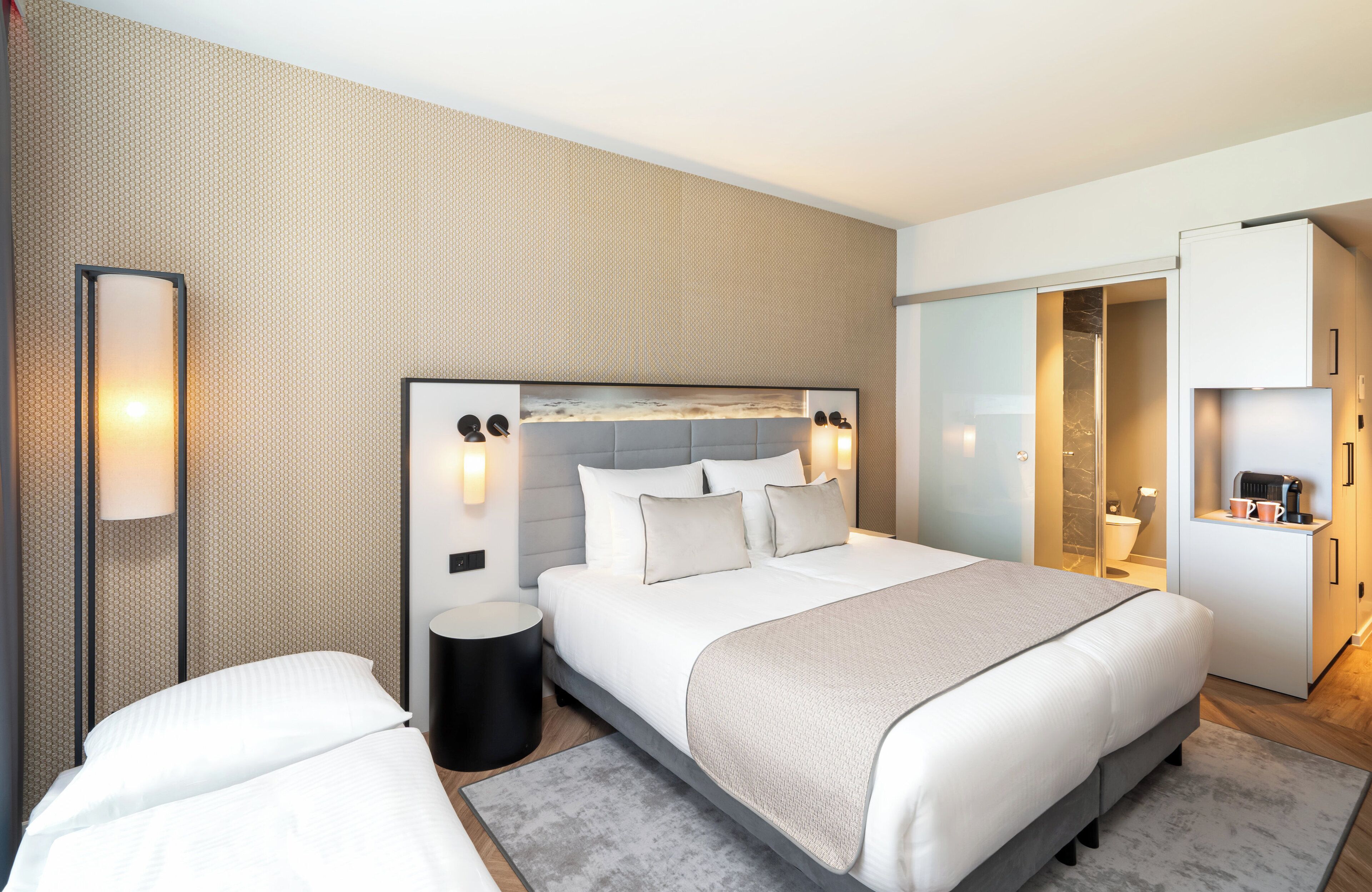 Quarto de hotel moderno com uma cama grande, uma mesa de cabeceira, um guarda-roupa e um banheiro visível através de uma porta aberta.