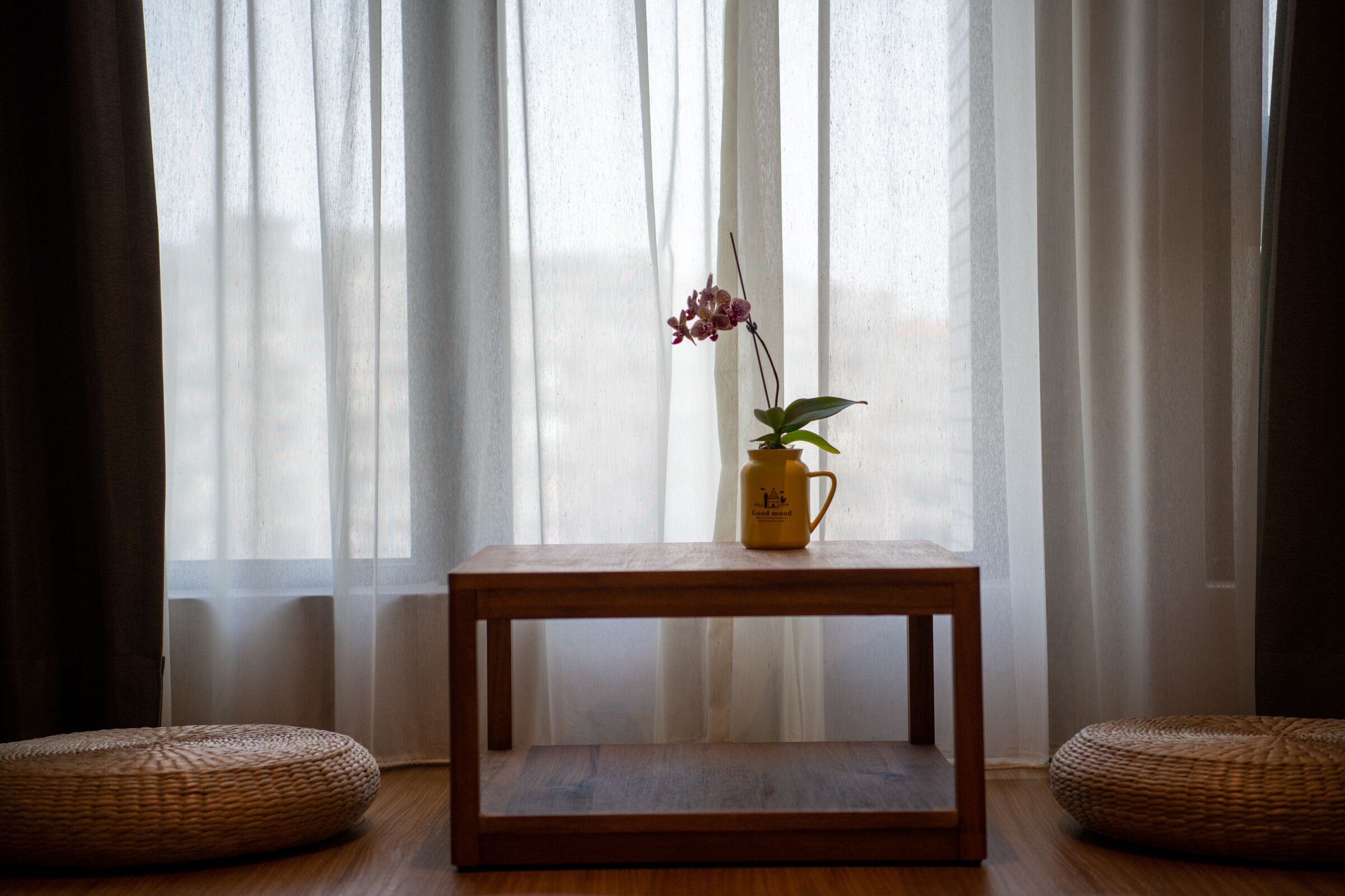 Uma mesa de madeira com uma orquídea em vaso e uma caneca amarela, colocada em frente a cortinas leves.