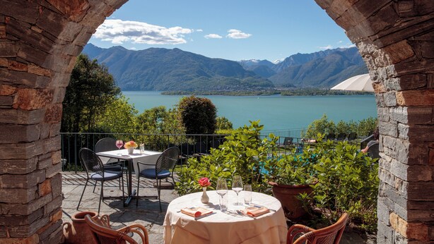 Um pátio externo com uma mesa posta para refeições, com vista para um lago e montanhas.