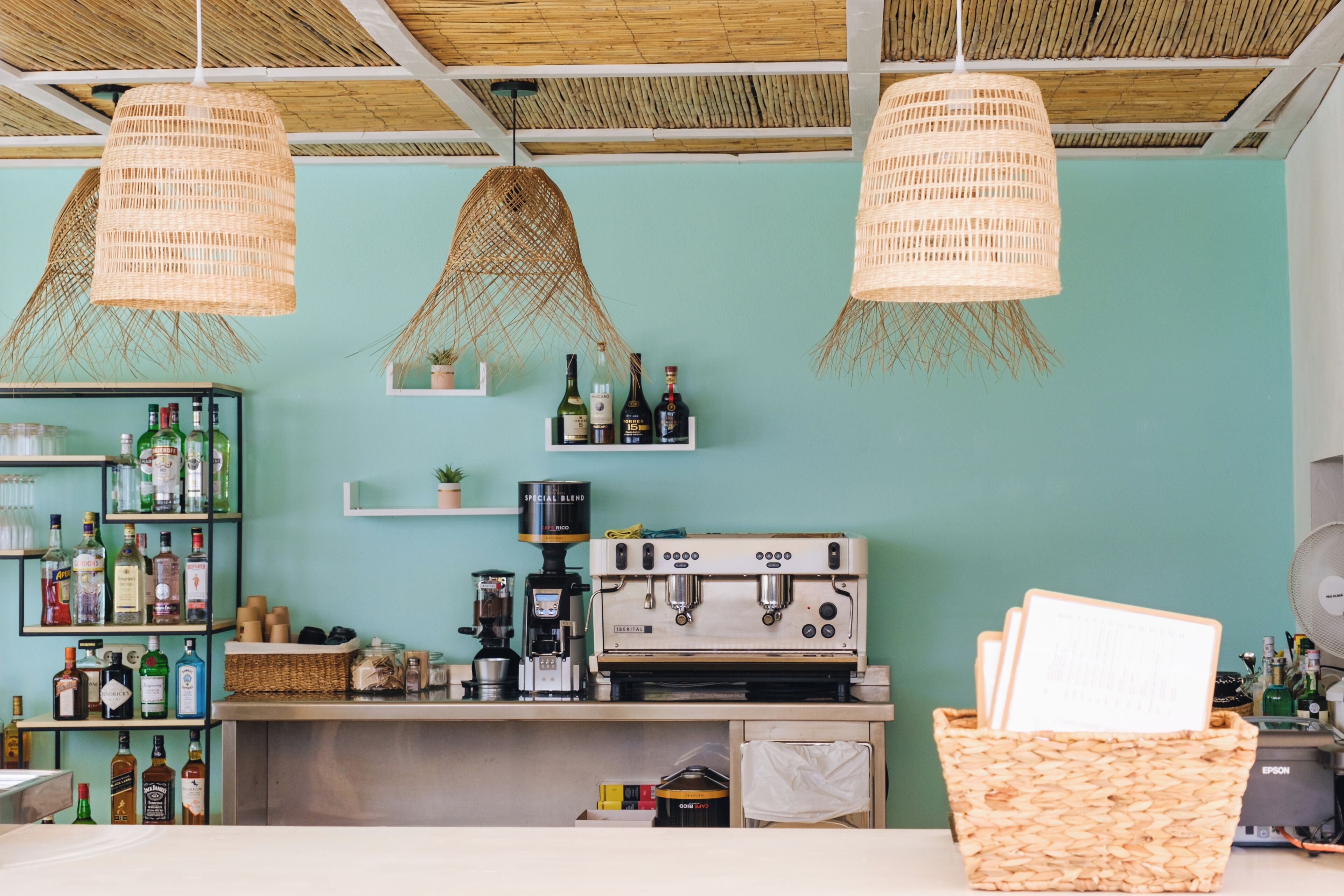 Interior de cafeteria com parede verde-turquesa, luminárias suspensas de vime, prateleiras com garrafas e uma máquina de espresso.
