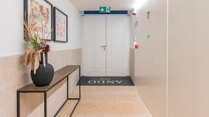 Um corredor com um balcão de recepção, obras de arte emolduradas, um vaso com plantas secas, uma porta com sinalização de saída de emergência e um tapete de entrada com a palavra 'ANDO'.