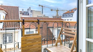 Varanda com cadeiras de madeira e grade de metal, com vista para paisagem urbana com telhados de terracota e guindastes de construção.