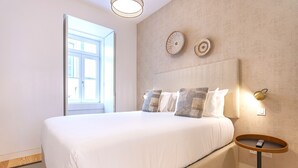 Cama bem arrumada com edredom branco e várias almofadas, mesa de cabeceira redonda de madeira com um controle remoto e uma luminária fixada na parede.