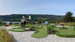 Um campo de golfe com uma grande escultura de alce, um lago e colinas ondulantes ao fundo.