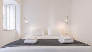 Cama bem arrumada com lençóis brancos e duas toalhas dobradas, um cobertor cinza texturizado e dois apliques de luz na parede.