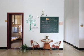 Um saguão de hotel com uma porta de madeira, uma cama de solteiro, uma mesa redonda com cadeiras e um quadro negro com texto e um enfeite de árvore.