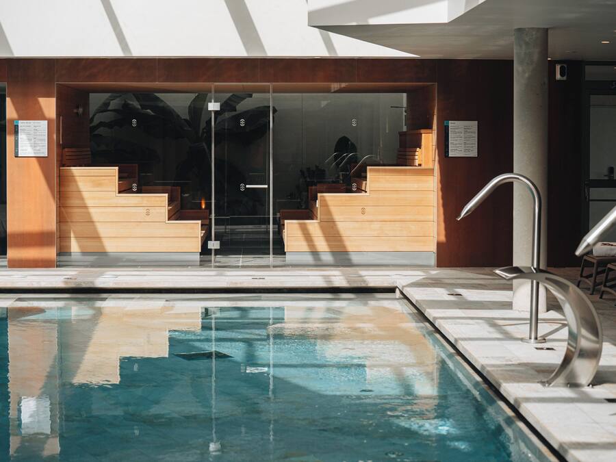 Piscina interna com água cristalina, escadas de madeira que levam a uma porta de vidro e corrimão de metal.