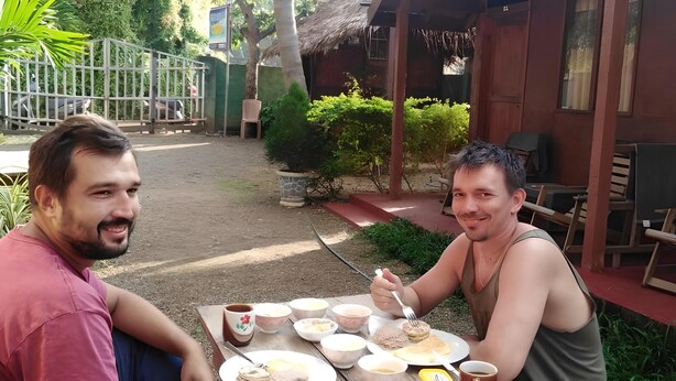 Duas pessoas jantando ao ar livre em uma mesa de madeira, com tigelas de comida e uma xícara.