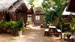 Uma área externa rústica com cabanas de palha, bancos de madeira e plantas em vasos.