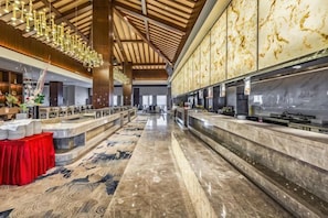 Um hall de hotel espaçoso, com teto alto, piso de mármore e um balcão de recepção central.
