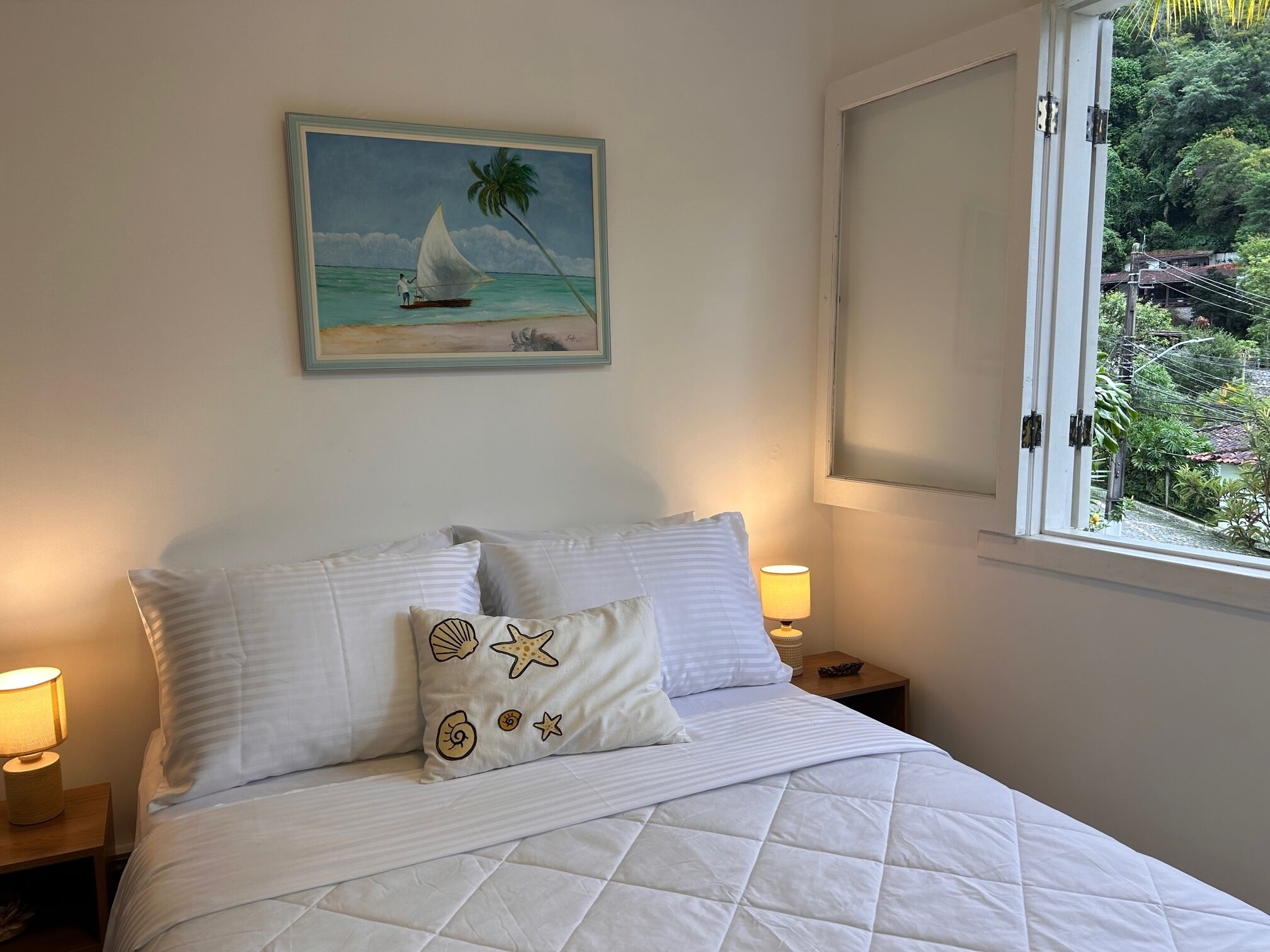 Quarto com cama, almofadas, mesa de cabeceira, abajur, pintura de um veleiro e janela com vista para vegetação.