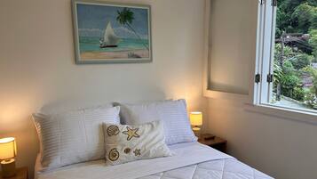 Quarto com cama, almofadas, mesa de cabeceira, abajur, pintura de um veleiro e janela com vista para vegetação.