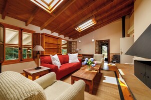 Uma sala de estar acolhedora com um sofá vermelho, uma poltrona bege e uma mesa de centro de madeira. Há uma lareira e uma janela grande com vista para o verde.